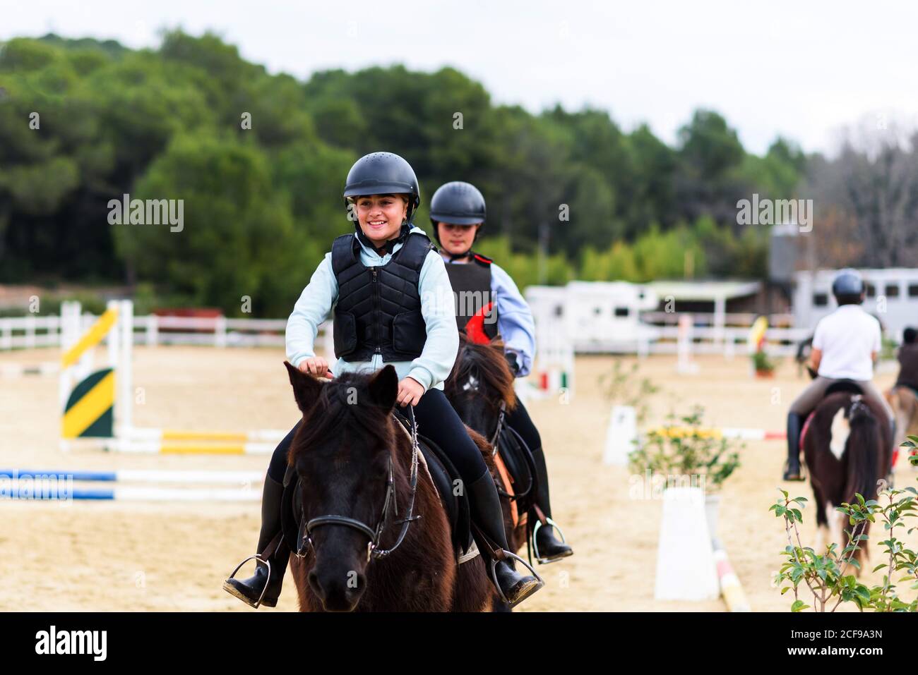 Happy teen jockeys in helmets riding obedient horses on sandy dressage ...
