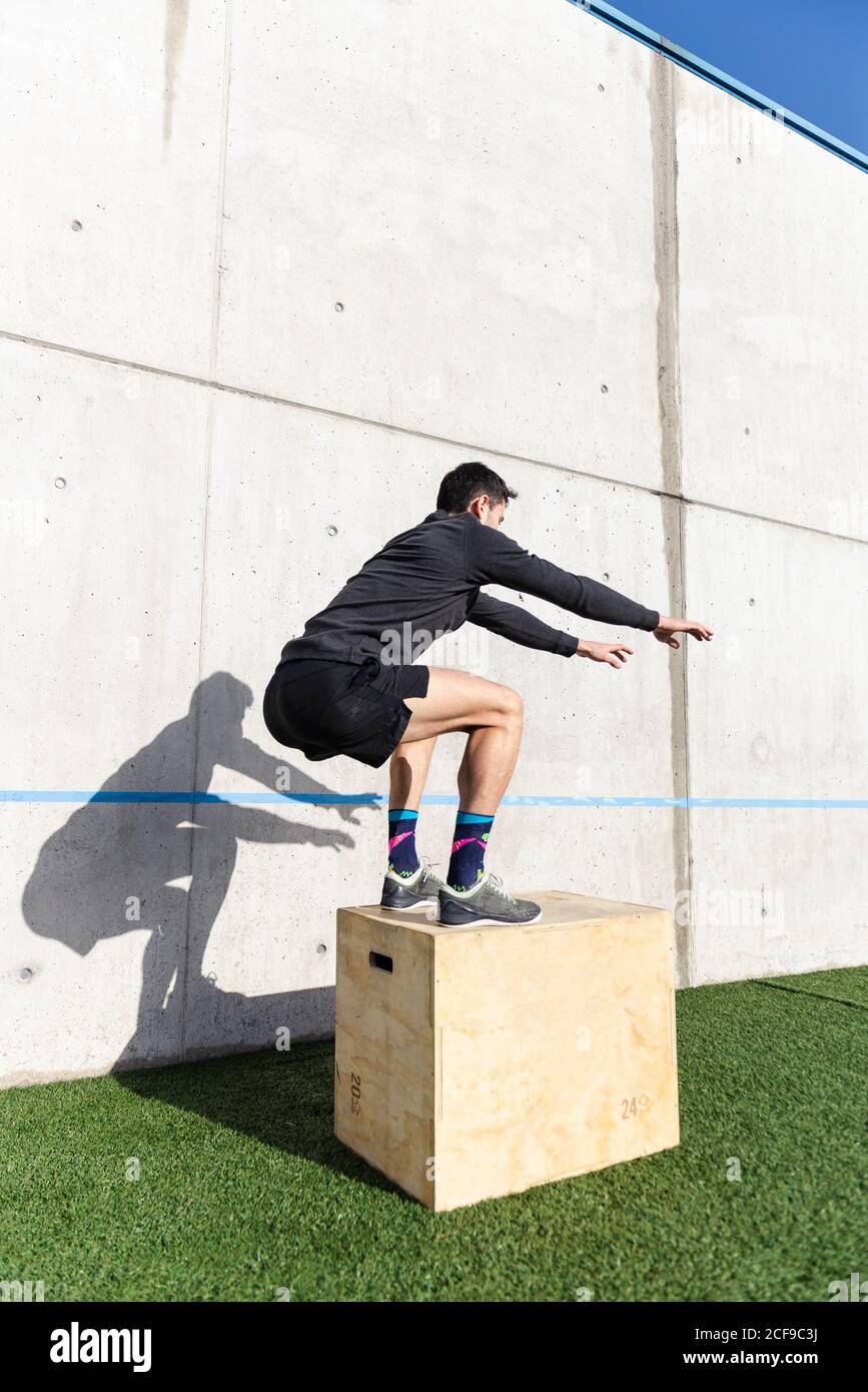 Side view of young athletic male jumping on box near grey wall in sunny ...
