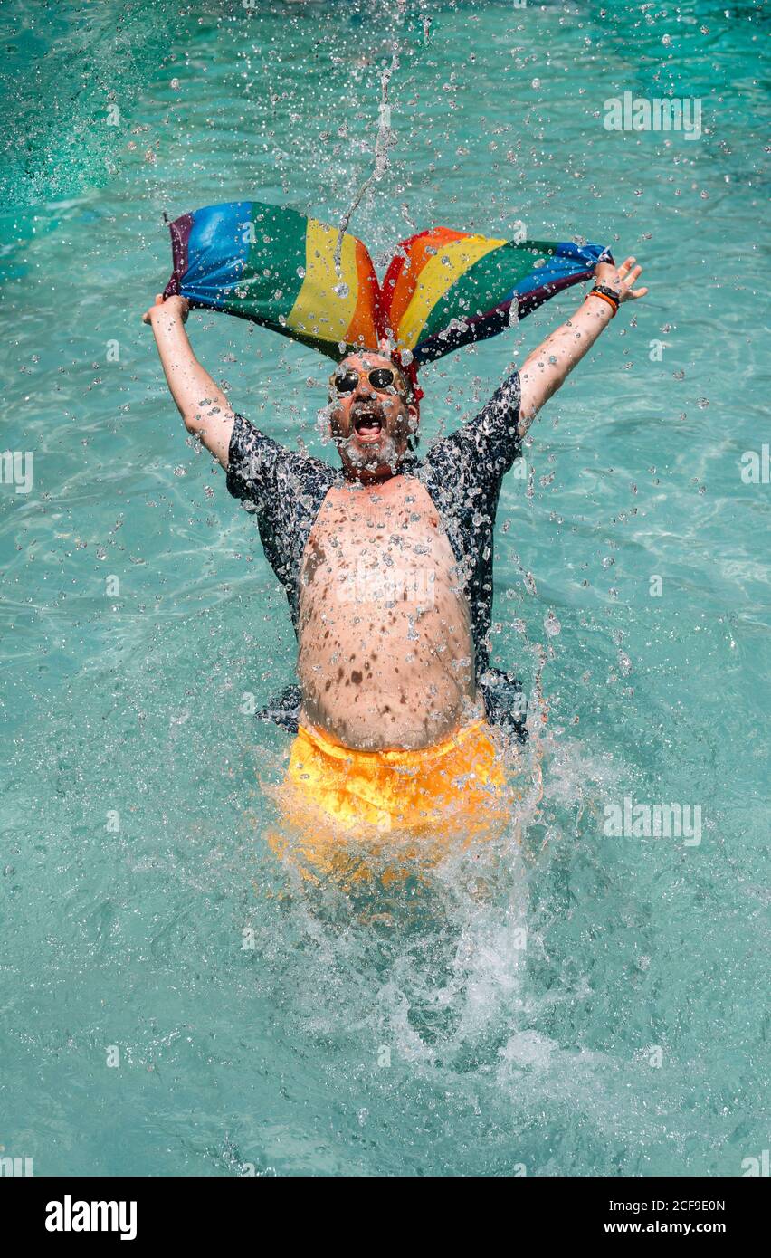 Cheerful man in swimwear screaming and waving LGBT flag while splashing ...
