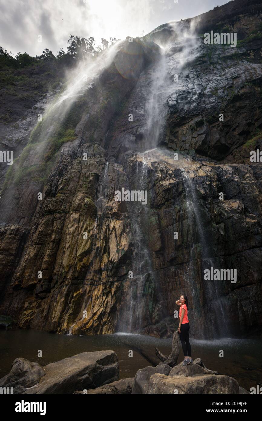 Foamy strong waterfall streaming from rocky mountain before small Woman ...
