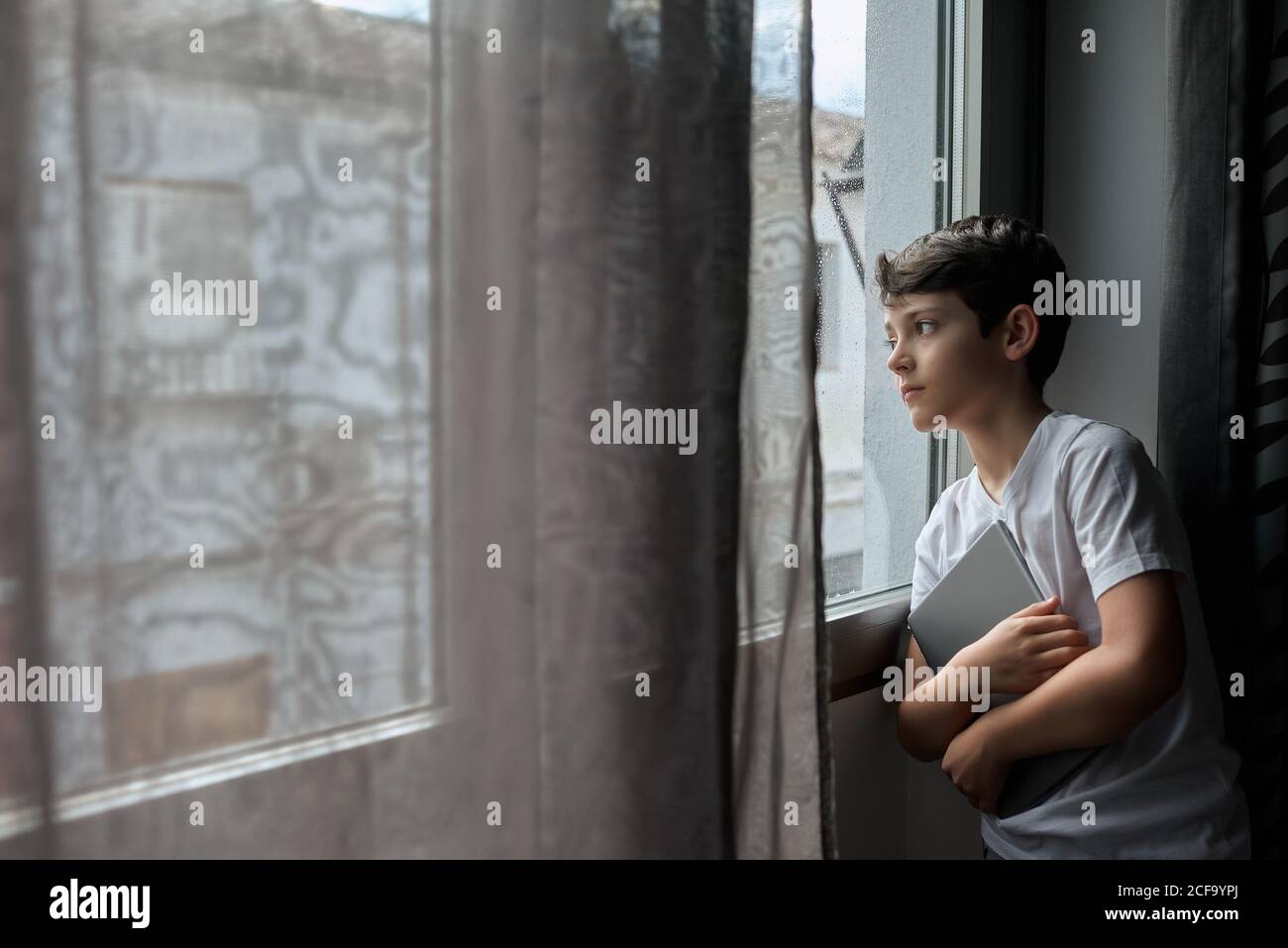 Thoughtful child in casual t shirt standing with tablet and leaning on ...