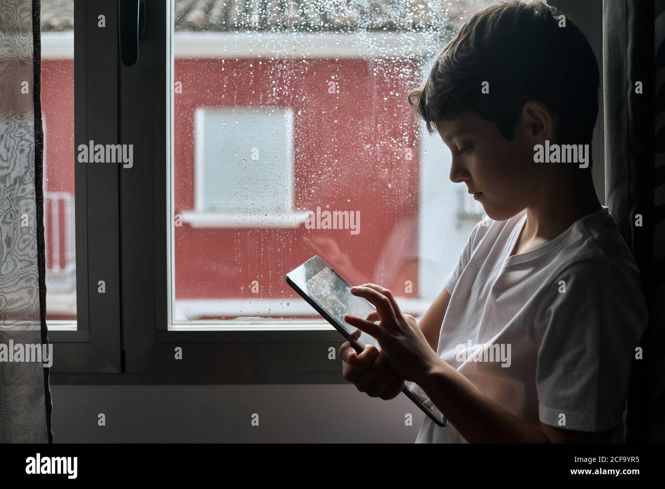 Thoughtful child in casual t shirt standing with tablet and leaning on ...