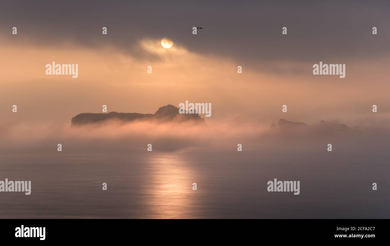 Calm surface of ocean and rough cliff under thick fog with bright sun ...