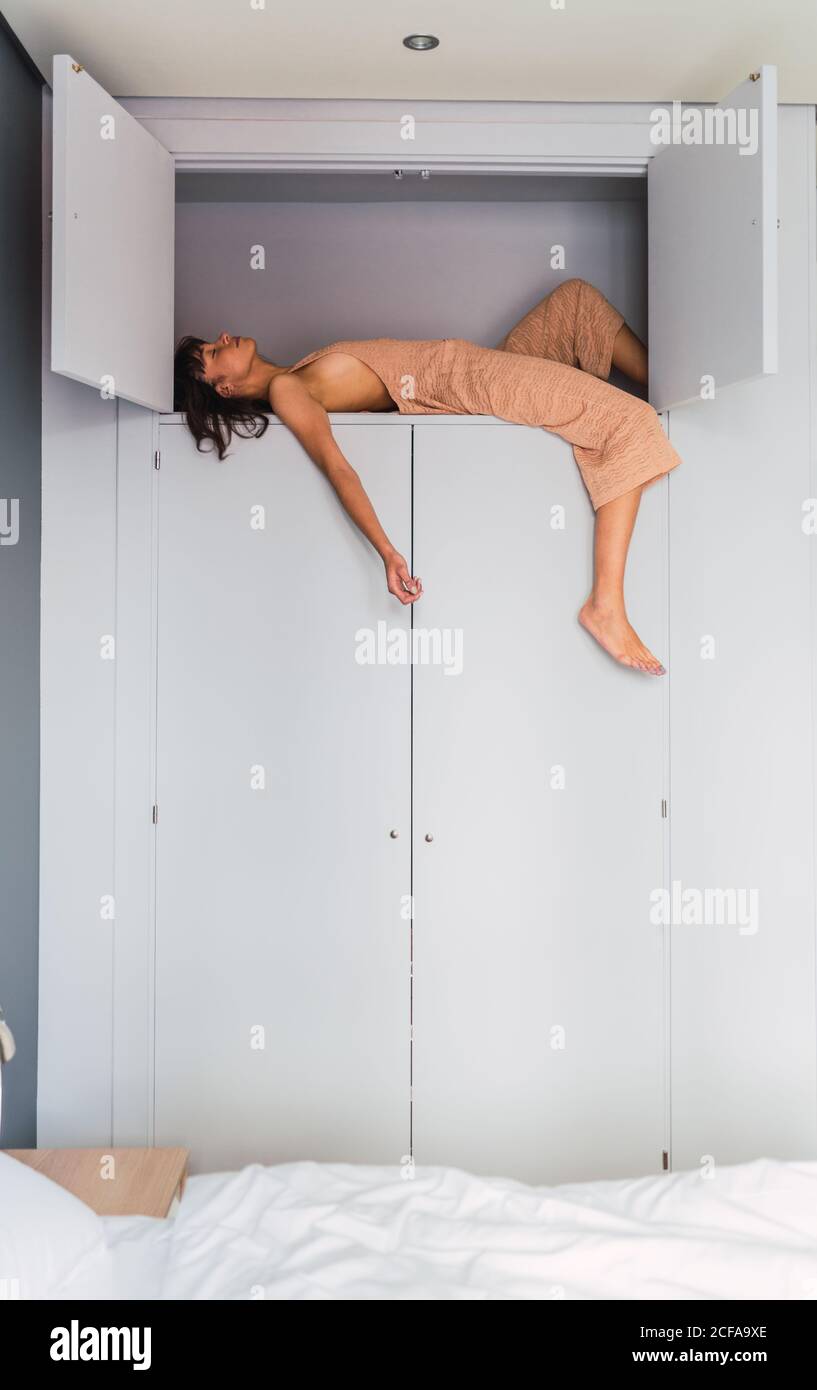 Side view of young Woman sleeping on high shelf of wardrobe near bed in ...