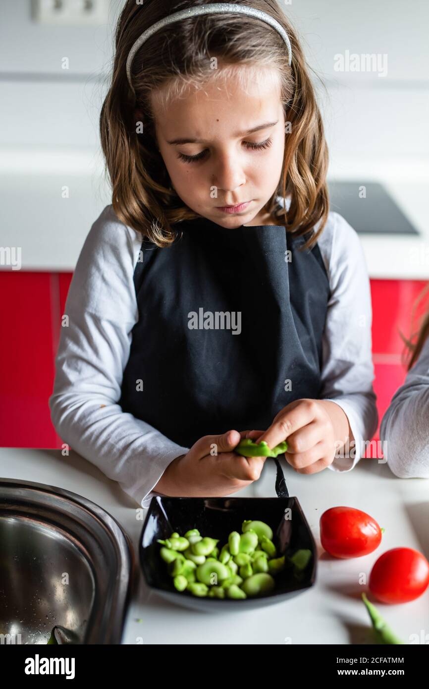 Little girl peeling ripe beans while cooking healthy salad in kitchen ...