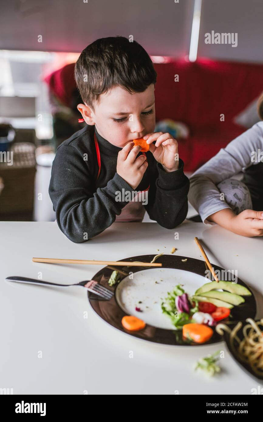boy eating vegetarian cutlets and vegetables while sitting at table at ...