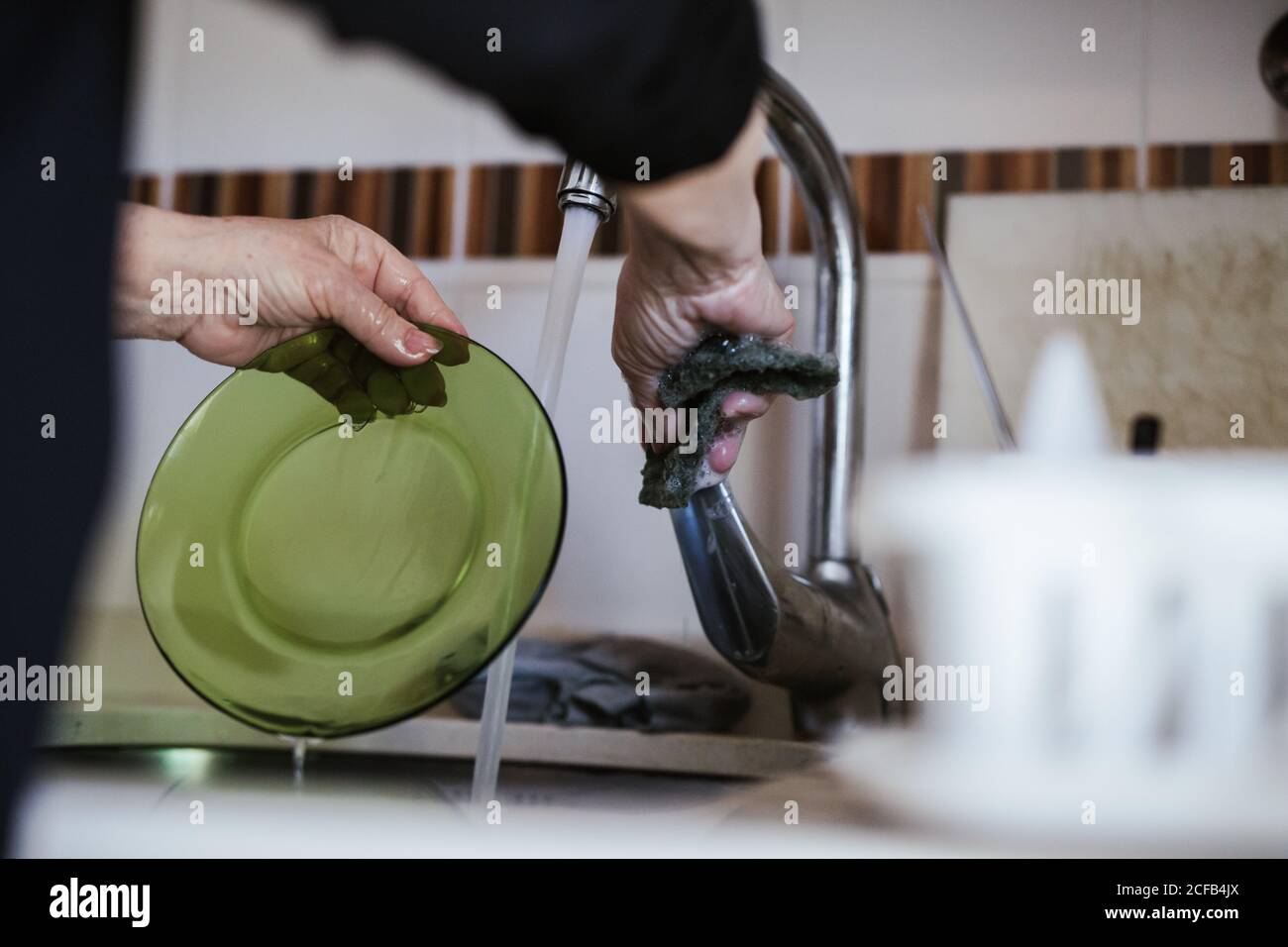 Back View Of Elderly Female Standing Near Sink And Washing Dish With Soap In Kitchen At Home