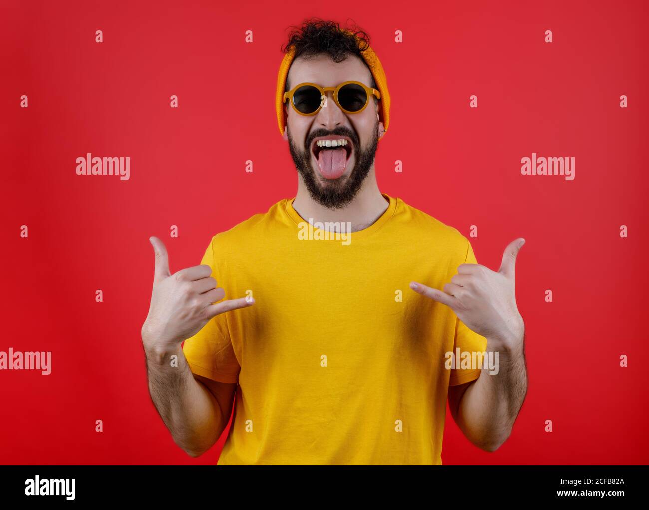 Bearded trendy man in yellow outfit showing tongue and gesturing shaka ...
