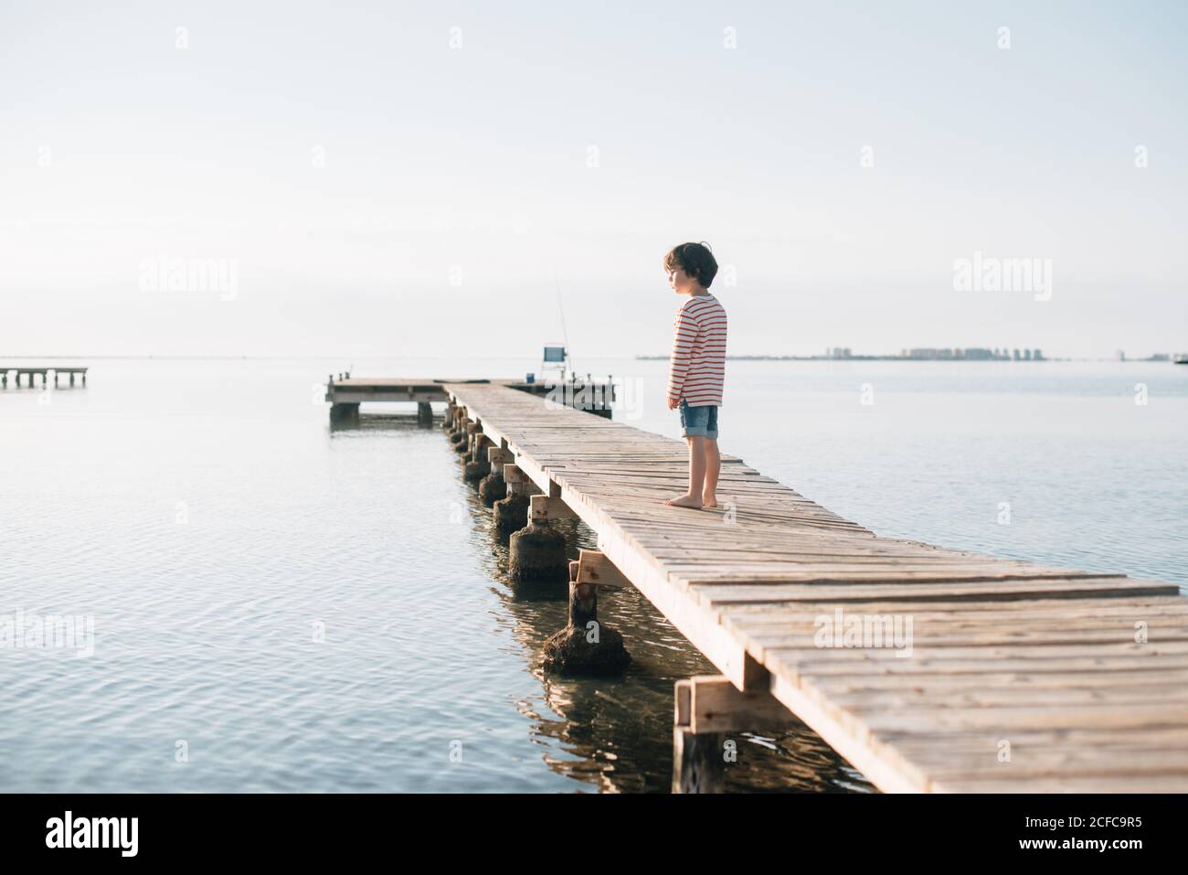 Back view of little boy standing on long wooden pier looking dreamily ...