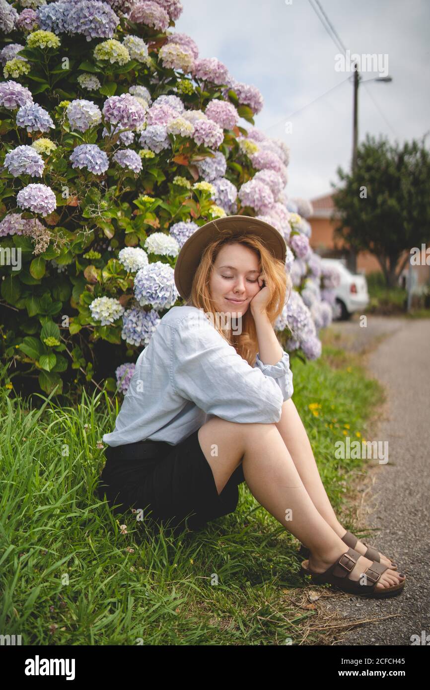 Smiling ginger carefree female closed eyes sitting among large bushes ...