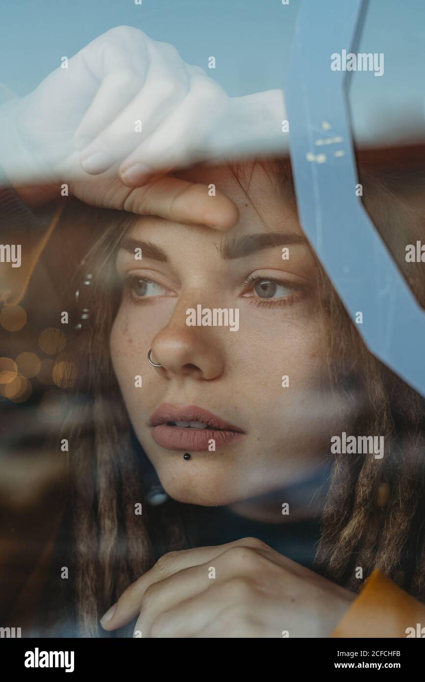 Pensive sad young female with dreadlocks leaning against window glass ...