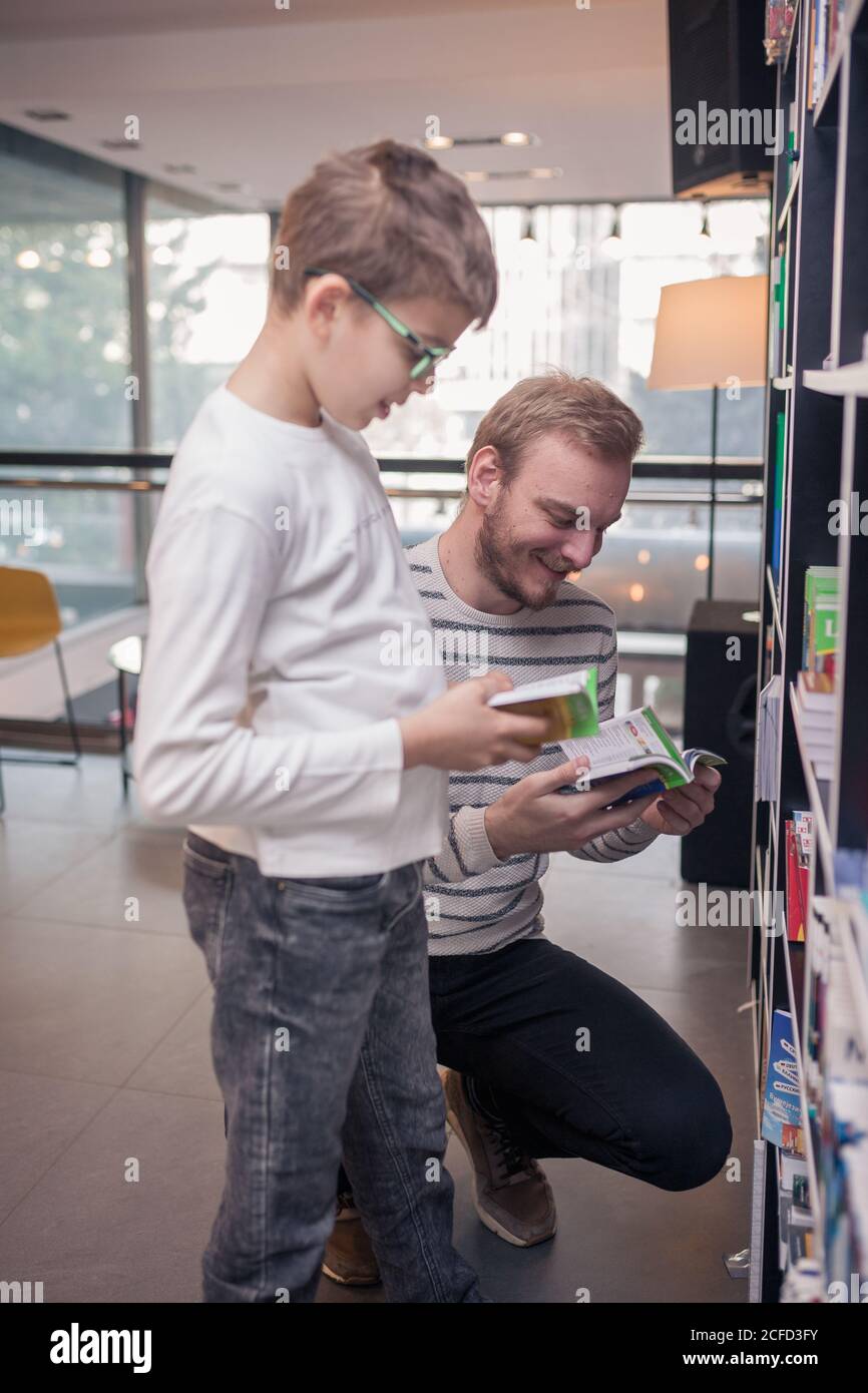 Son and father reading books in front of bookshelf in library Stock ...