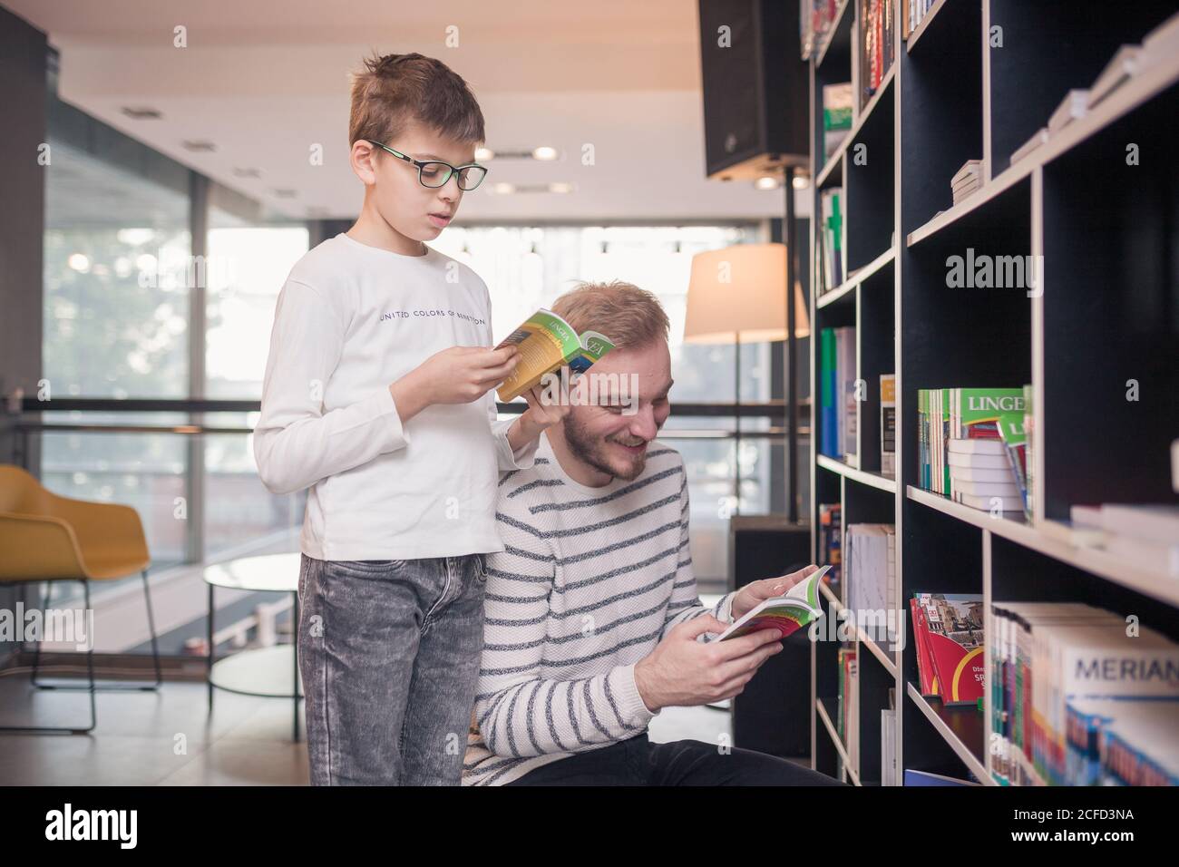 Son and father reading books and smiling in front of bookshelf in ...