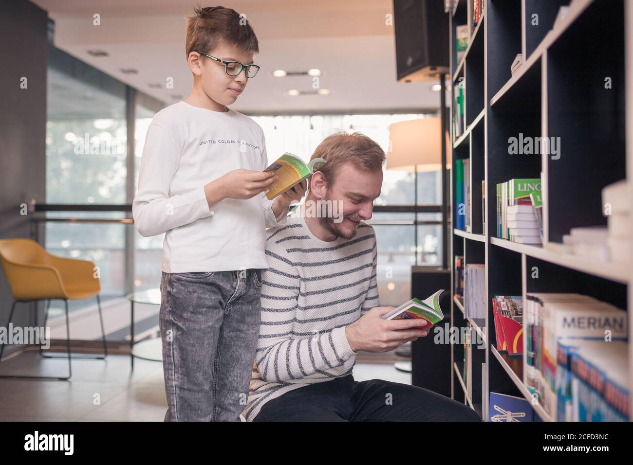 Son and father reading books in front of bookshelf in library Stock ...