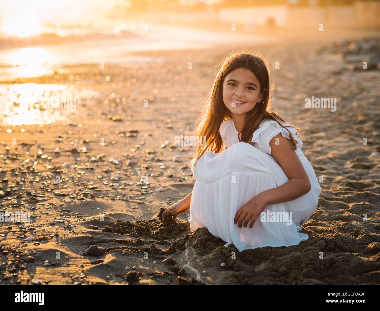 Cute pensive female kid in white dress playing with sand on seaside in ...