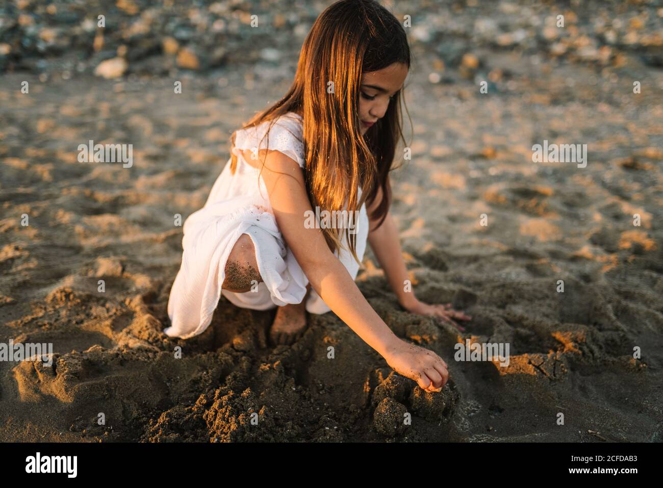 Cute pensive female kid in white dress playing with sand on seaside in ...