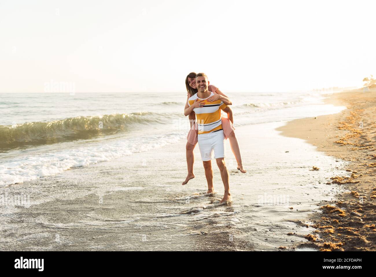 Full length cheerful adult male giving piggyback ride to smiling female ...