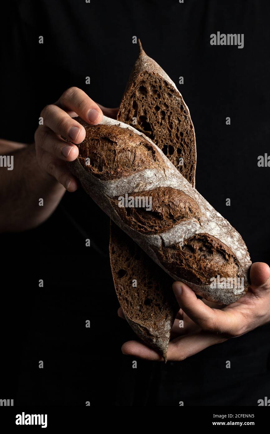 Crop male baker in apron holding cut in half loaf of fresh healthy ...