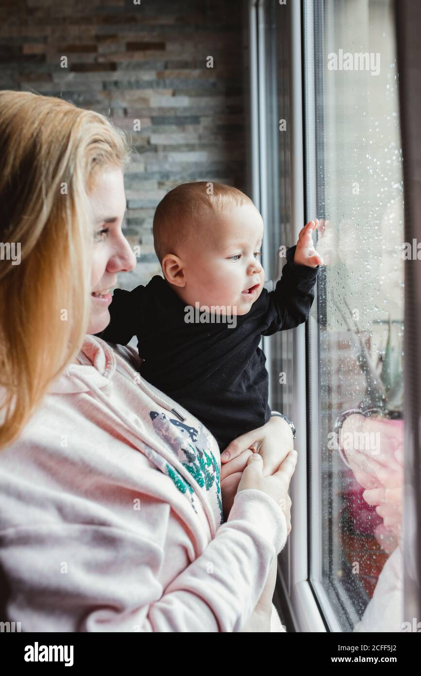Young Woman carrying cute baby and looking out window on rainy day at ...