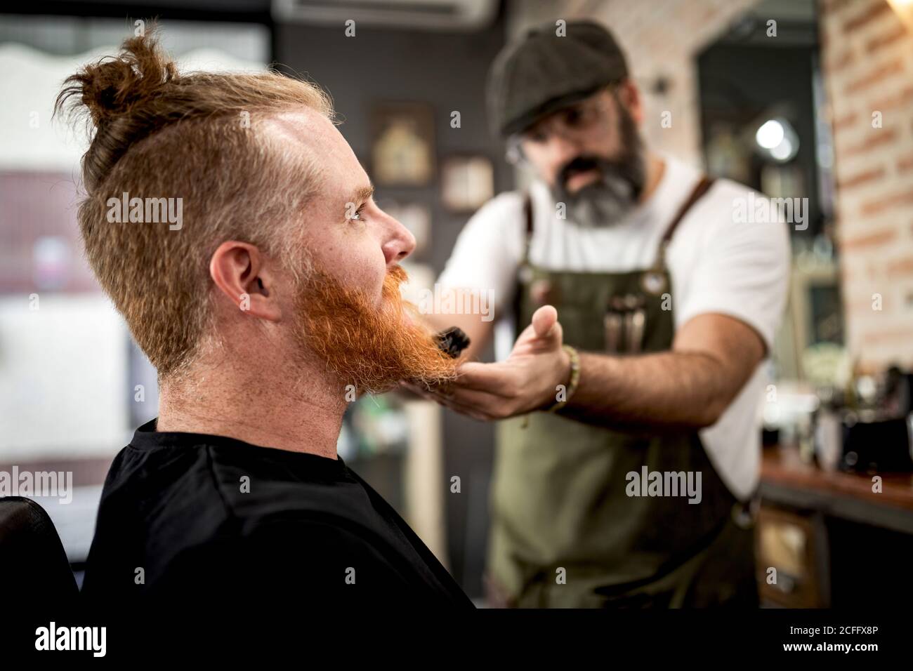 Barber with trimmer cutting beard of redhead man sitting in barbershop ...