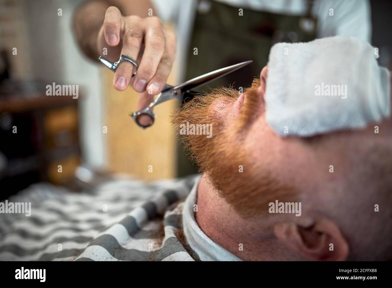 Cropped unrecognizable barber with scissors cutting beard of redhead ...