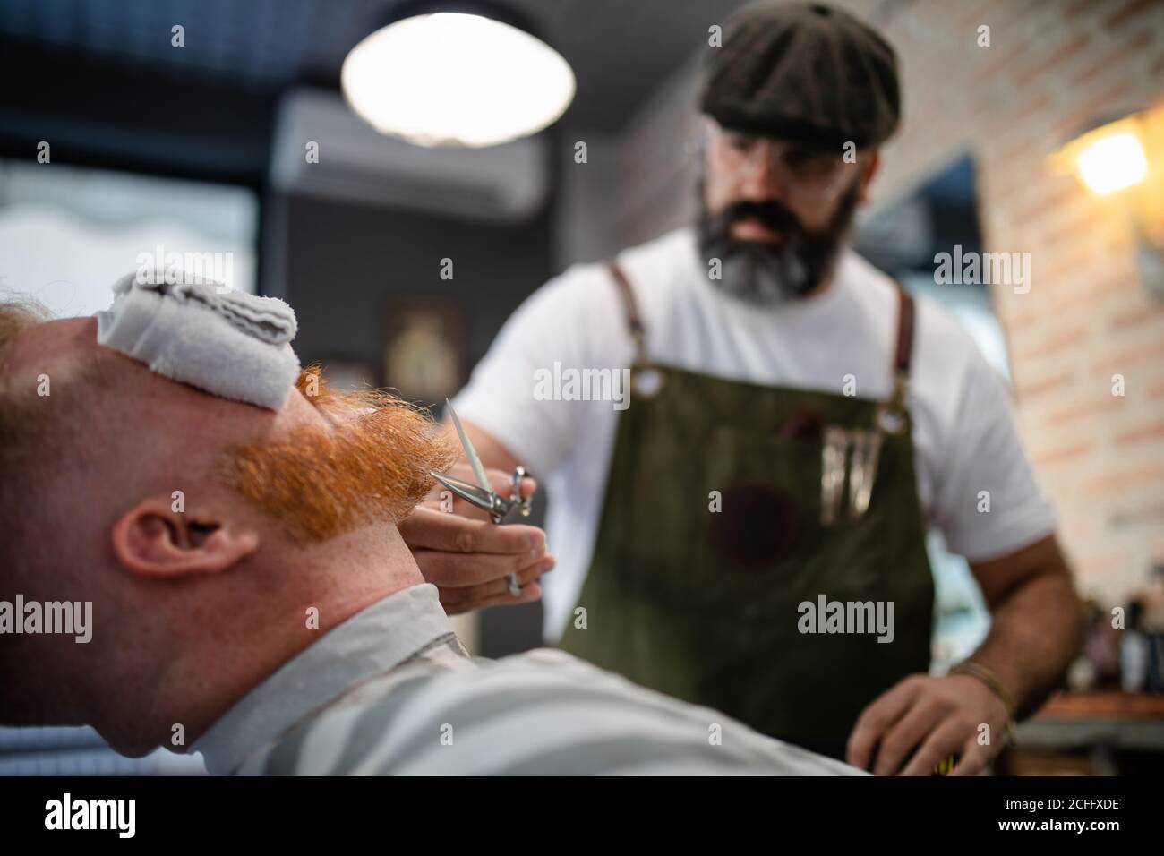 Blurred unrecognizable barber with scissor fixing redhead man beard ...