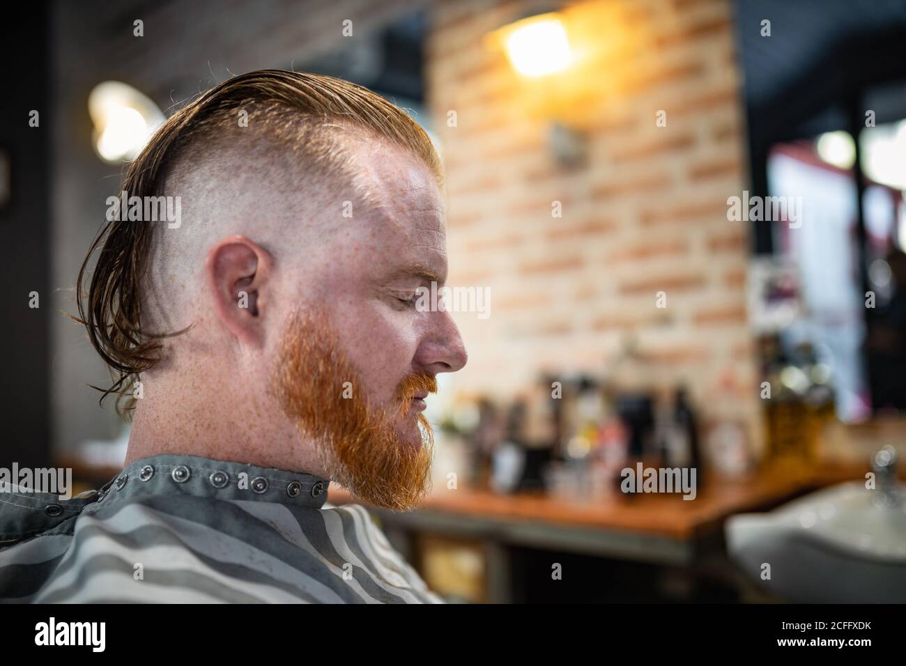 Side view of redhead man sitting in modern barbershop with closed eyes ...