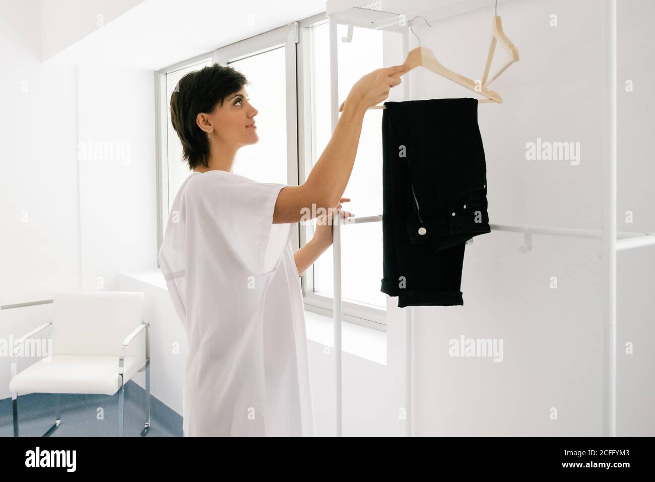 Side view of happy female in white robe hanging pants on rail while ...