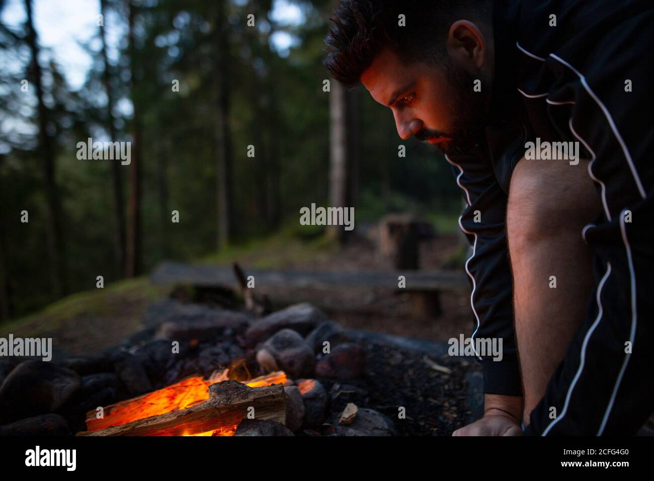 Side view of tranquil man sitting near campfire in woods and warming up ...