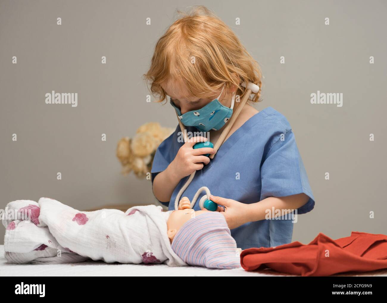 young child wearing medical PPE examines a baby doll by checking it's ...