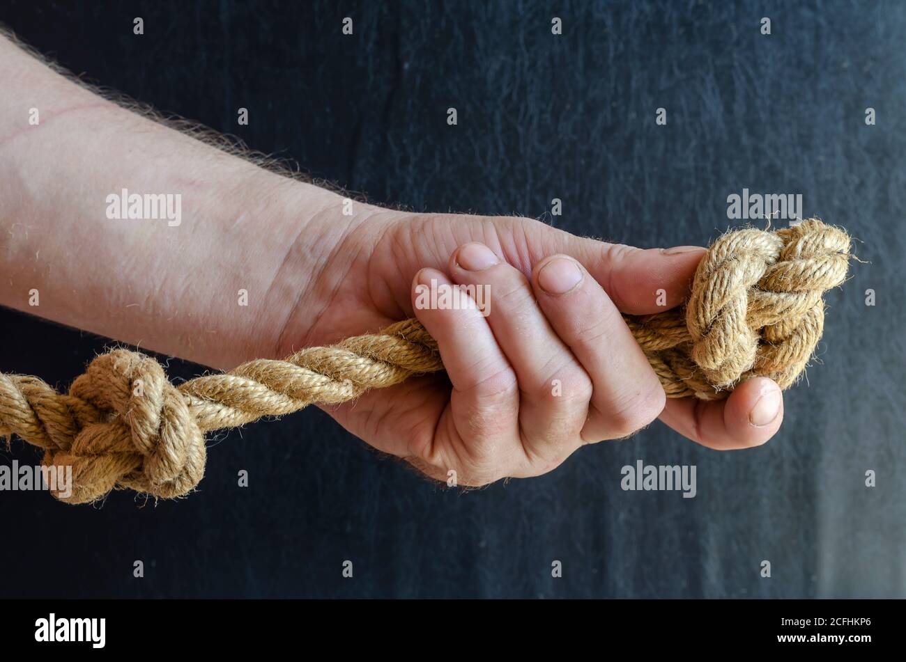 A man's hand holds a rope with a knotted knot on a dark background. A ...