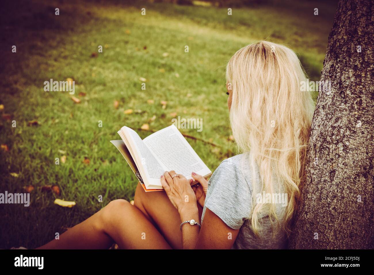Young blonde woman reading a book in a park, sitting on the grass and ...