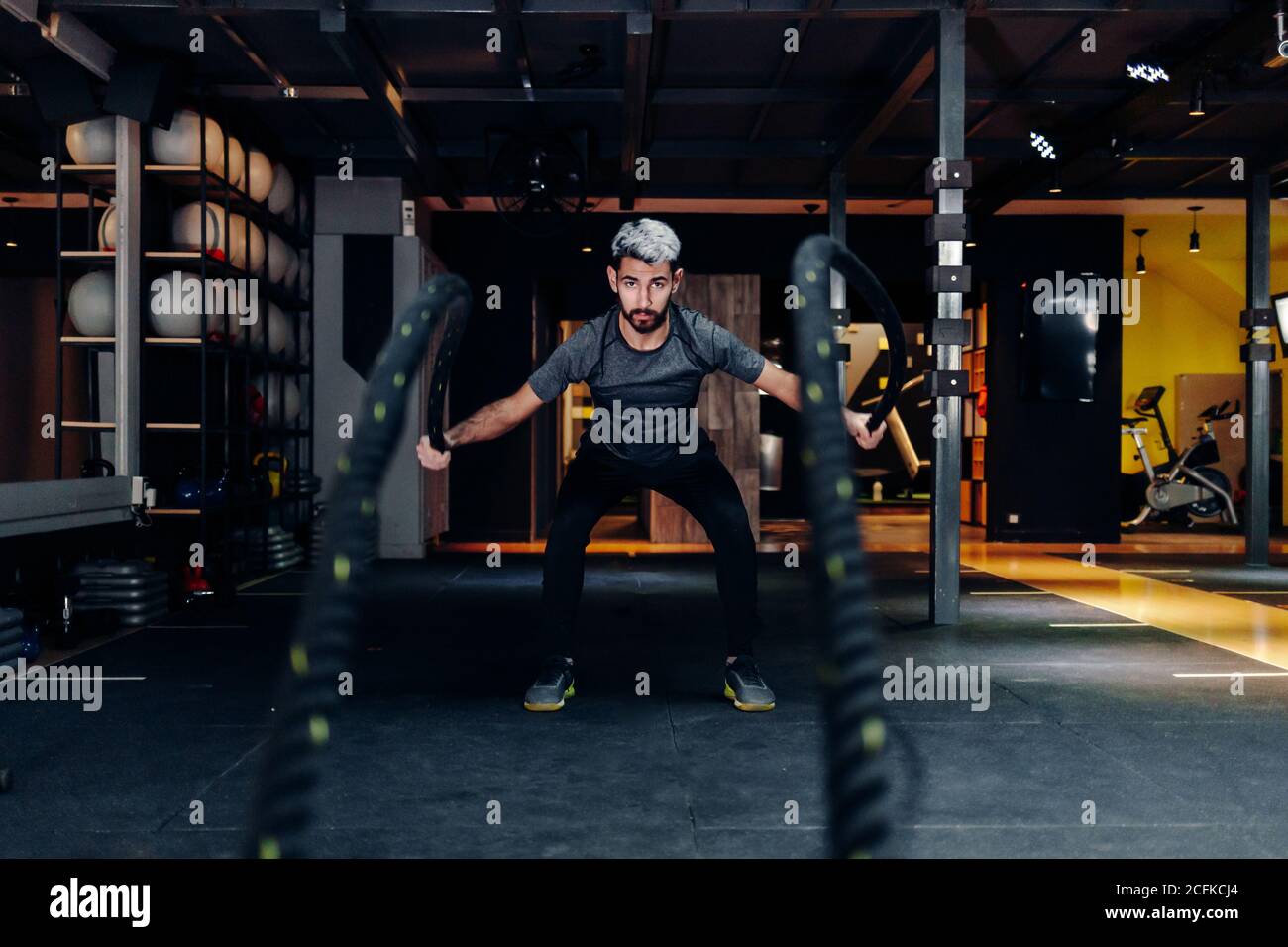 Focused determined young sporty man training with battle ropes during ...