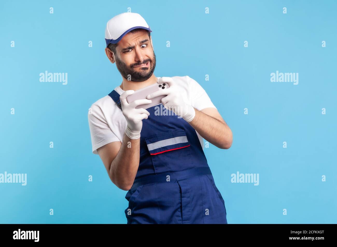 Funny surprised handyman in overalls and gloves holding mobile phone ...