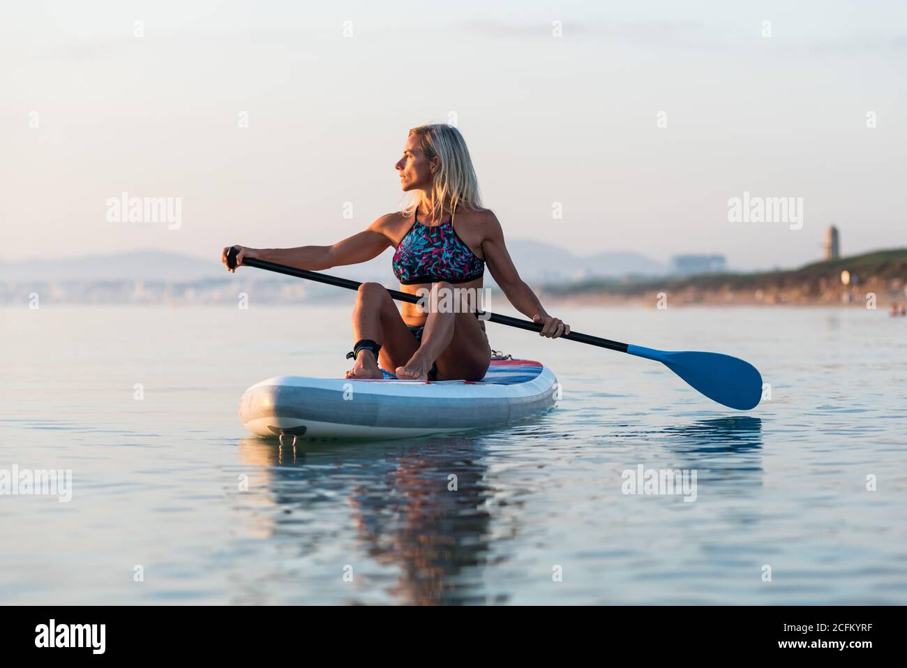 Side view of fit female surfer sitting on paddleboard and rowing ...