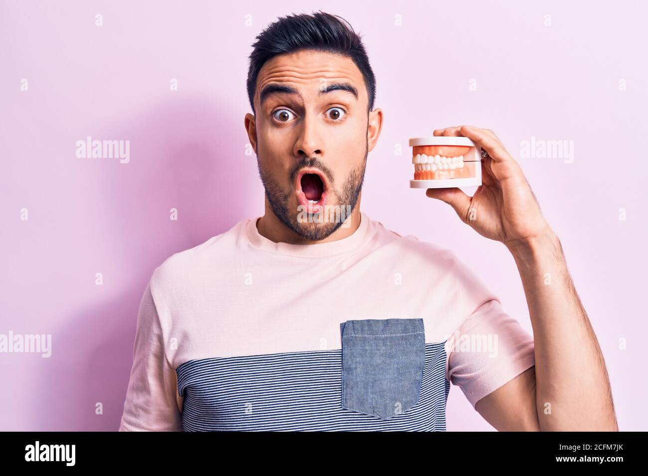 Young handsome man with beard holding denture teeth over isolated pink ...