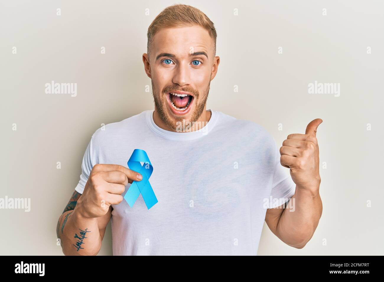 Young caucasian man holding blue ribbon pointing thumb up to the side ...