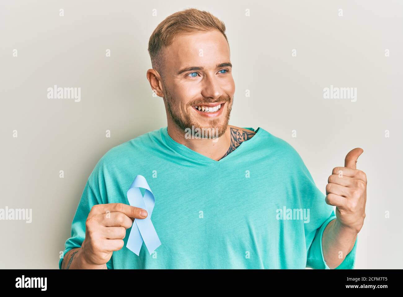 Young caucasian man holding blue ribbon pointing thumb up to the side ...