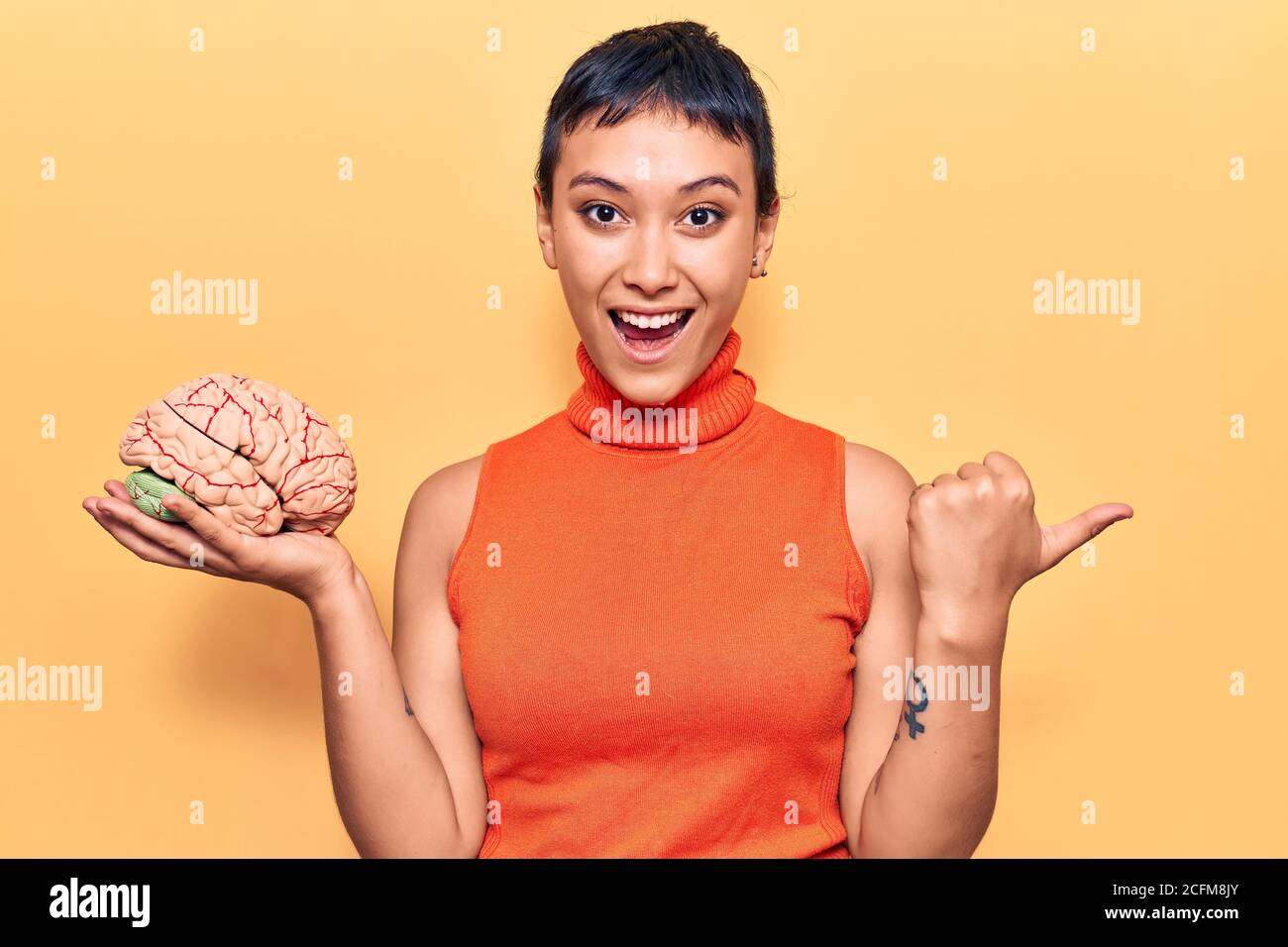 Young woman holding brain pointing thumb up to the side smiling happy ...