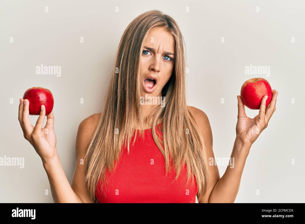 Young blonde woman holding red apples in shock face, looking skeptical ...