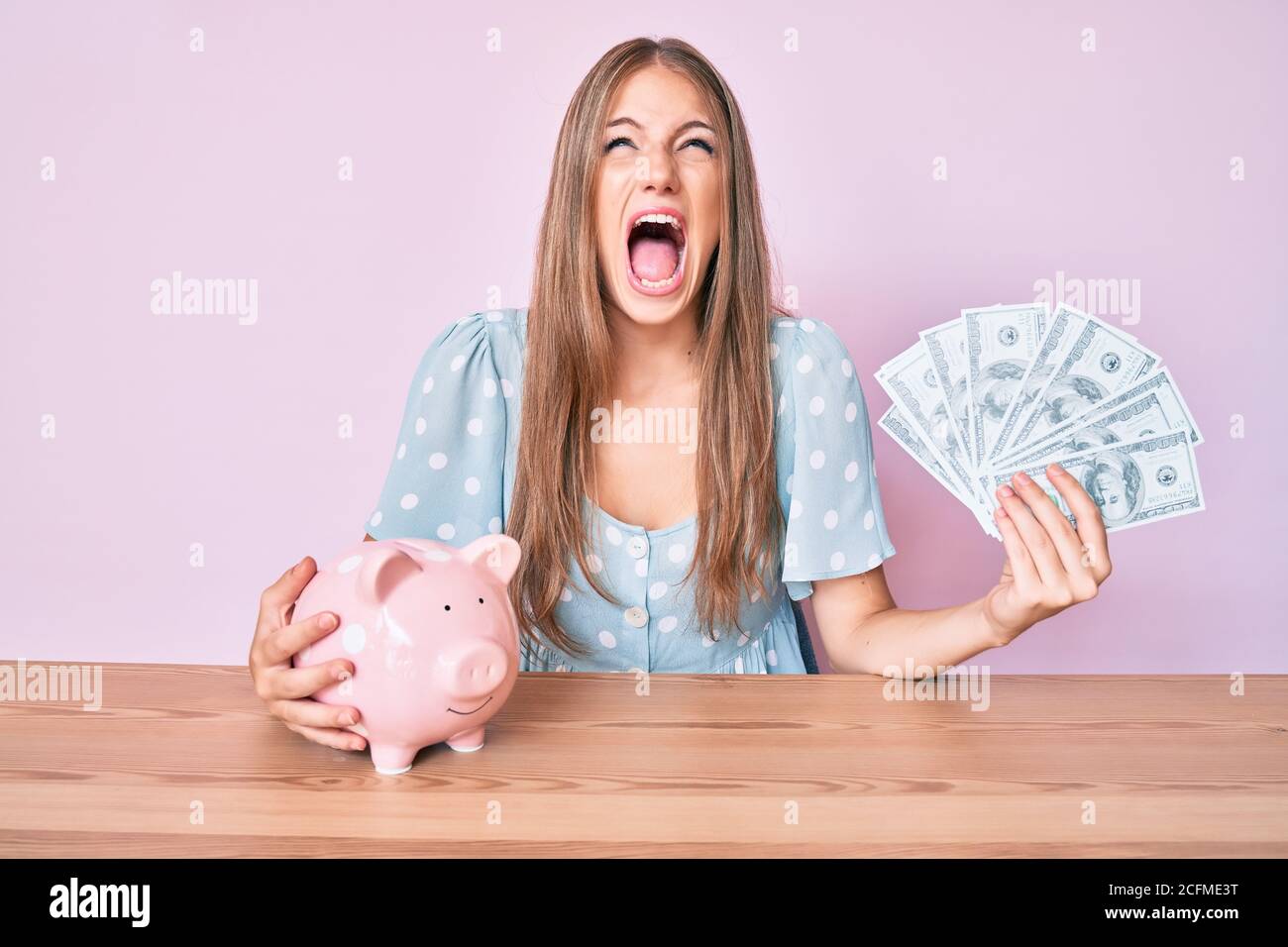 Young blonde girl holding dollars and piggy bank sitting on the table ...