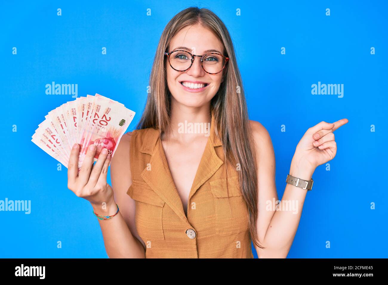 Young blonde girl holding israeli shekels smiling happy pointing with ...