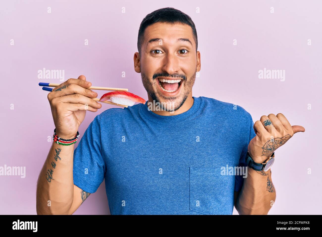 Handsome man with tattoos eating tuna sushi using chopsticks pointing ...
