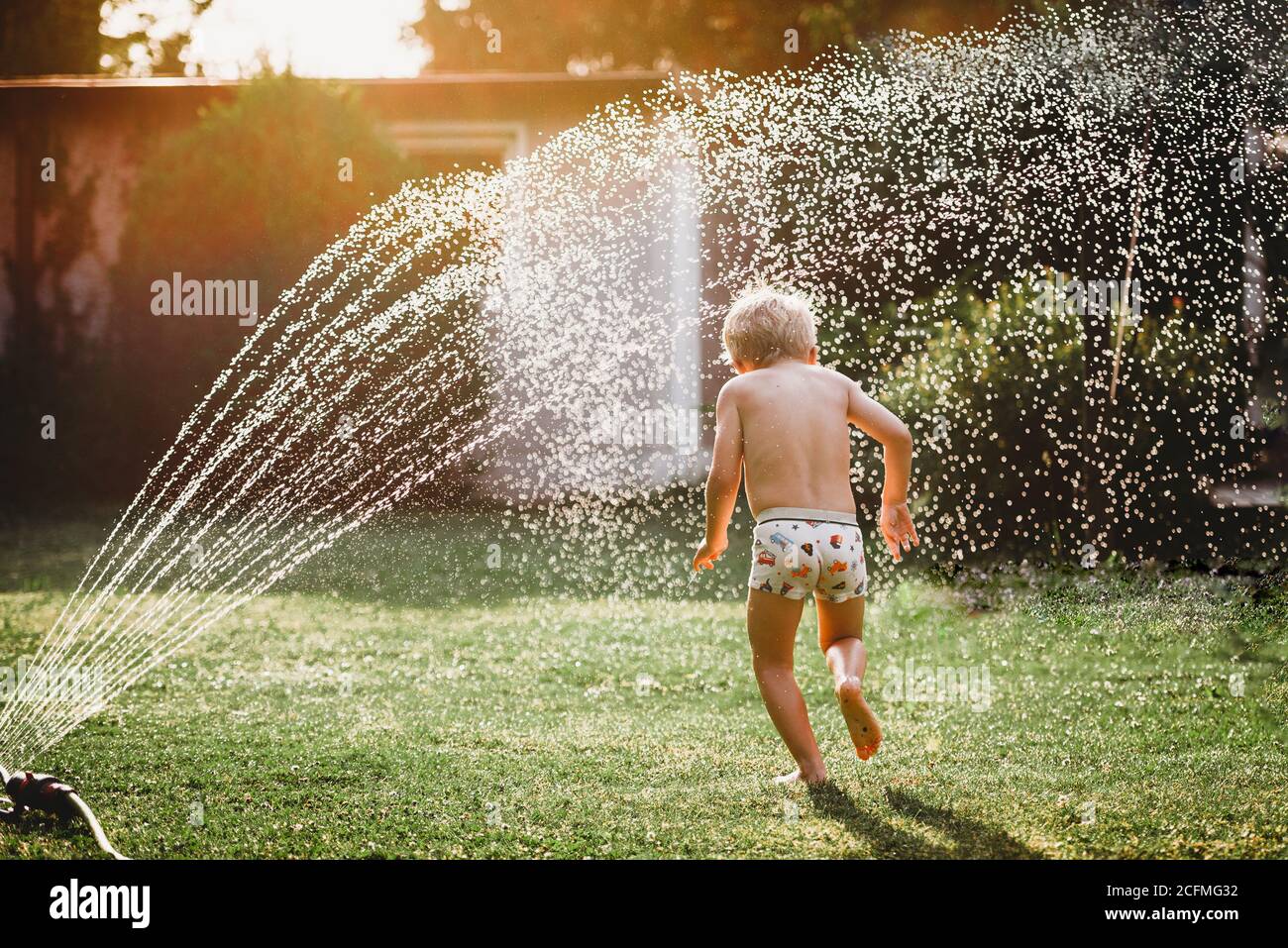 Young white boy running under the water from the sprinkler in garden ...