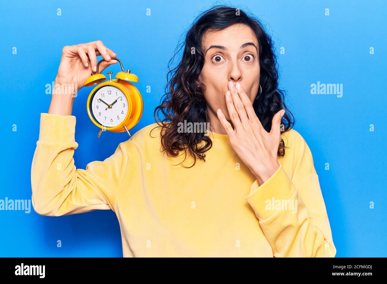 Young beautiful hispanic woman holding alarm clock covering mouth with ...