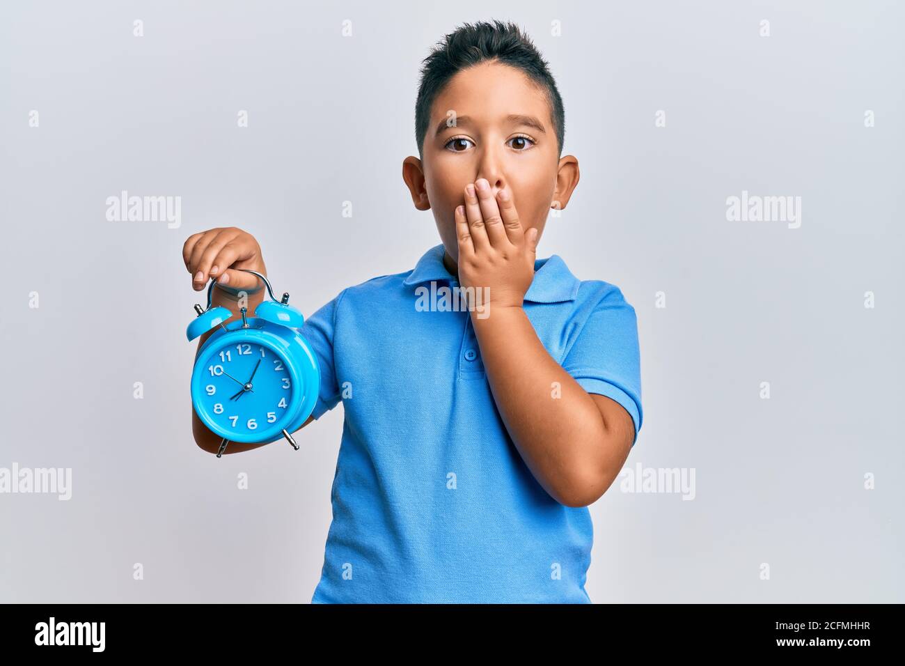 Little boy hispanic kid holding alarm clock covering mouth with hand ...