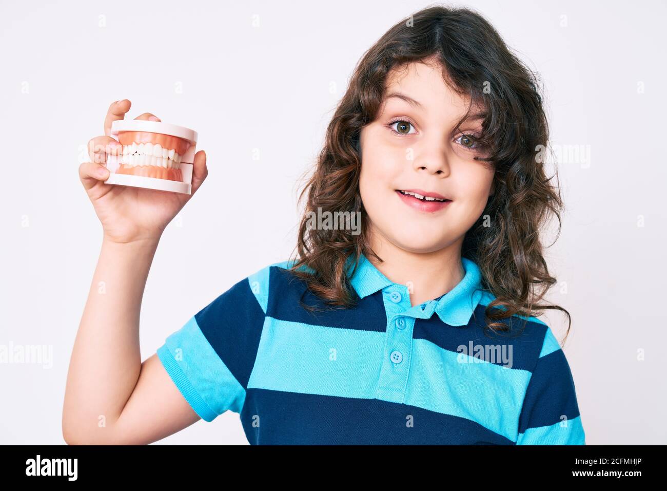 Cute hispanic child with long hair holding denture looking positive and ...