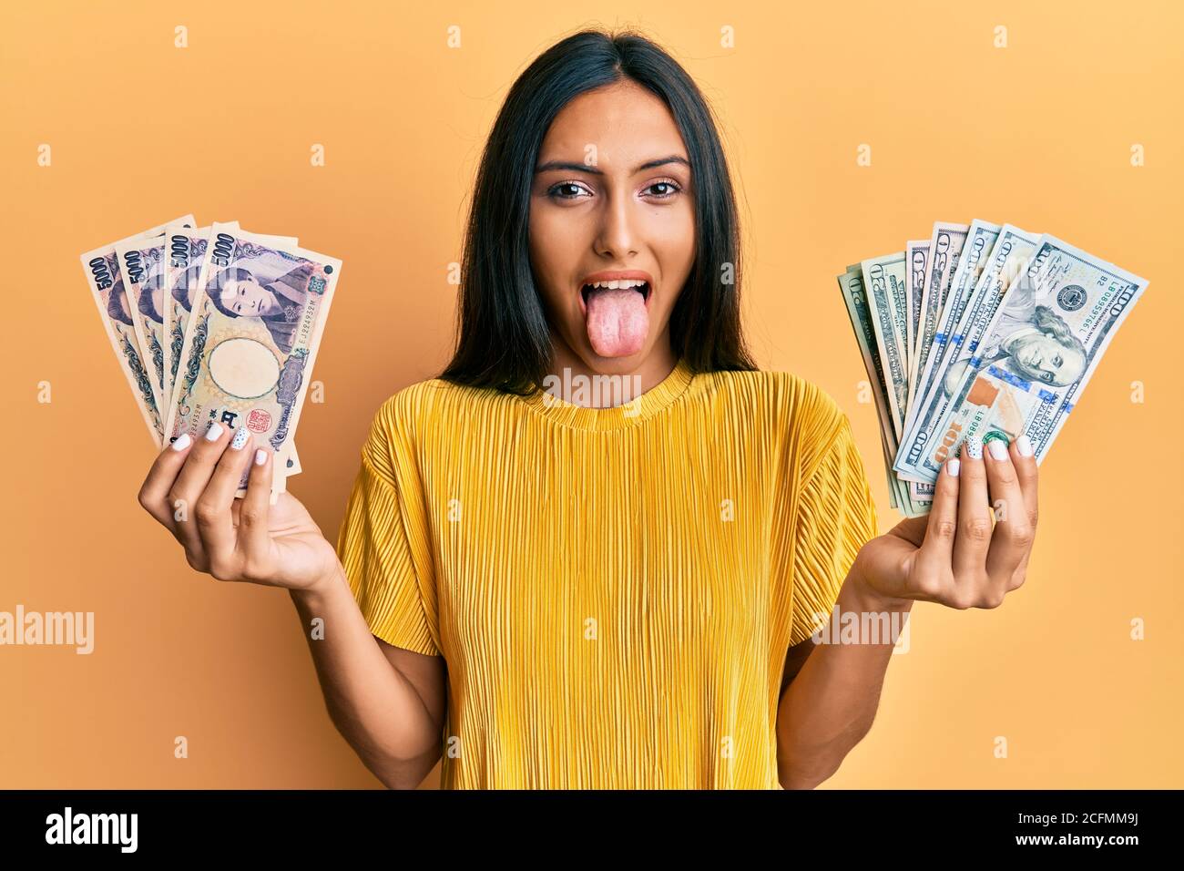 Young brunette woman holding bunch of dollars and yens sticking tongue ...