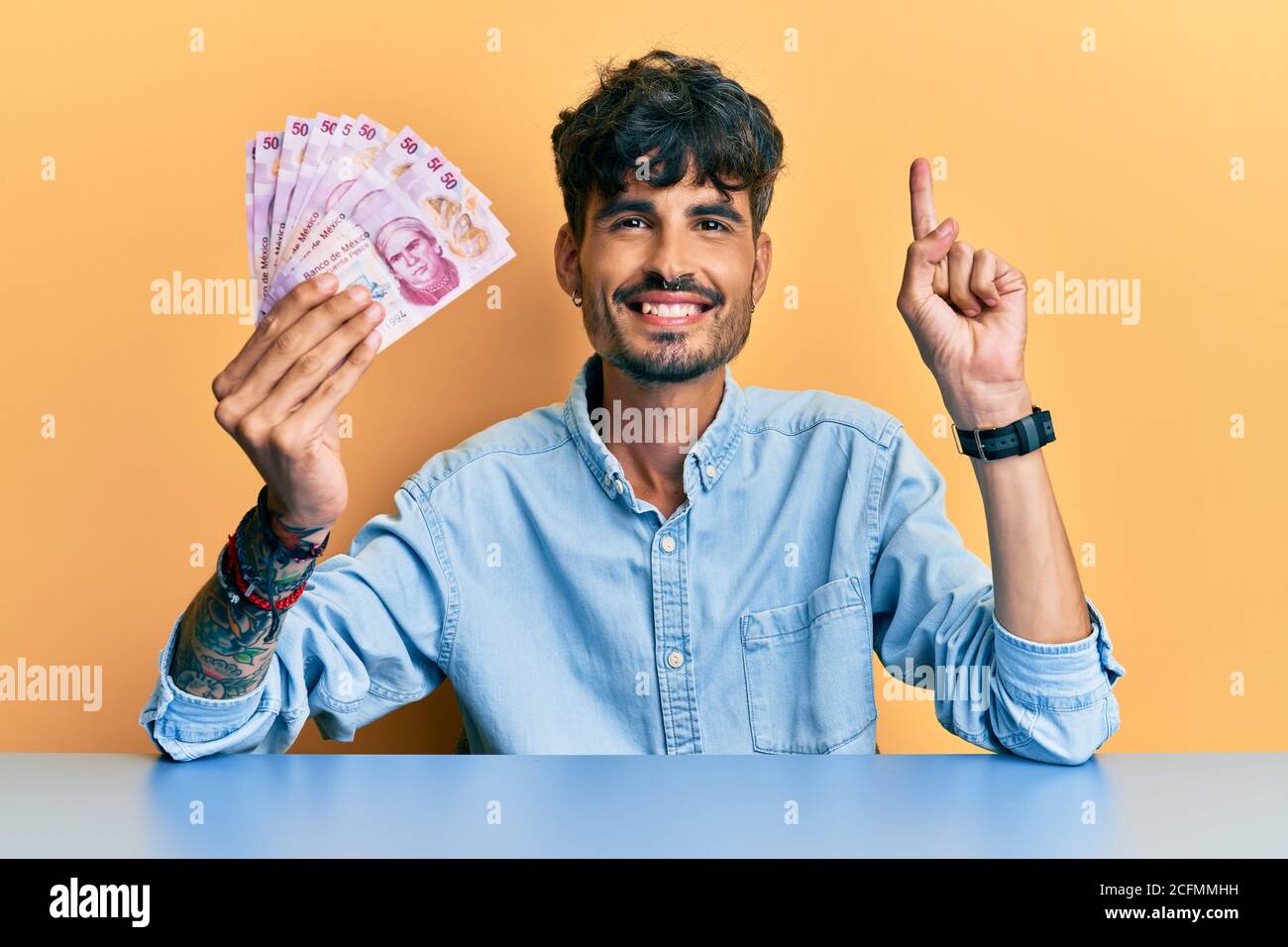 Young hispanic man holding mexican pesos sitting on the table smiling ...