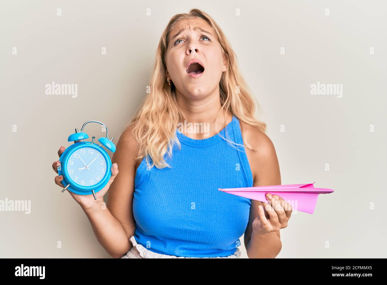 Young blonde girl holding paper plane and alarm clock angry and mad ...